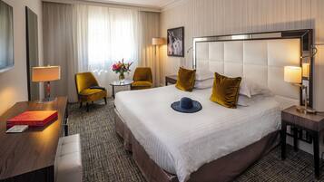 Quarto de hotel com uma cama grande, uma escrivaninha com um laptop, uma cadeira, uma mesa pequena com um vaso de flores e uma luminária.