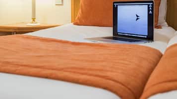 Uma cama bem arrumada com um cobertor laranja, um laptop sobre a cama e uma luminária em uma mesa de cabeceira.