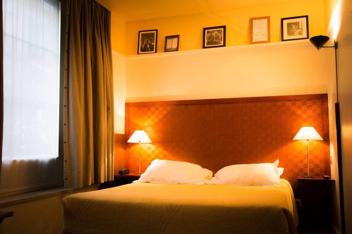 Quarto de hotel com uma cama grande, duas luminárias de cabeceira, uma janela com cortinas e quadros nas paredes.