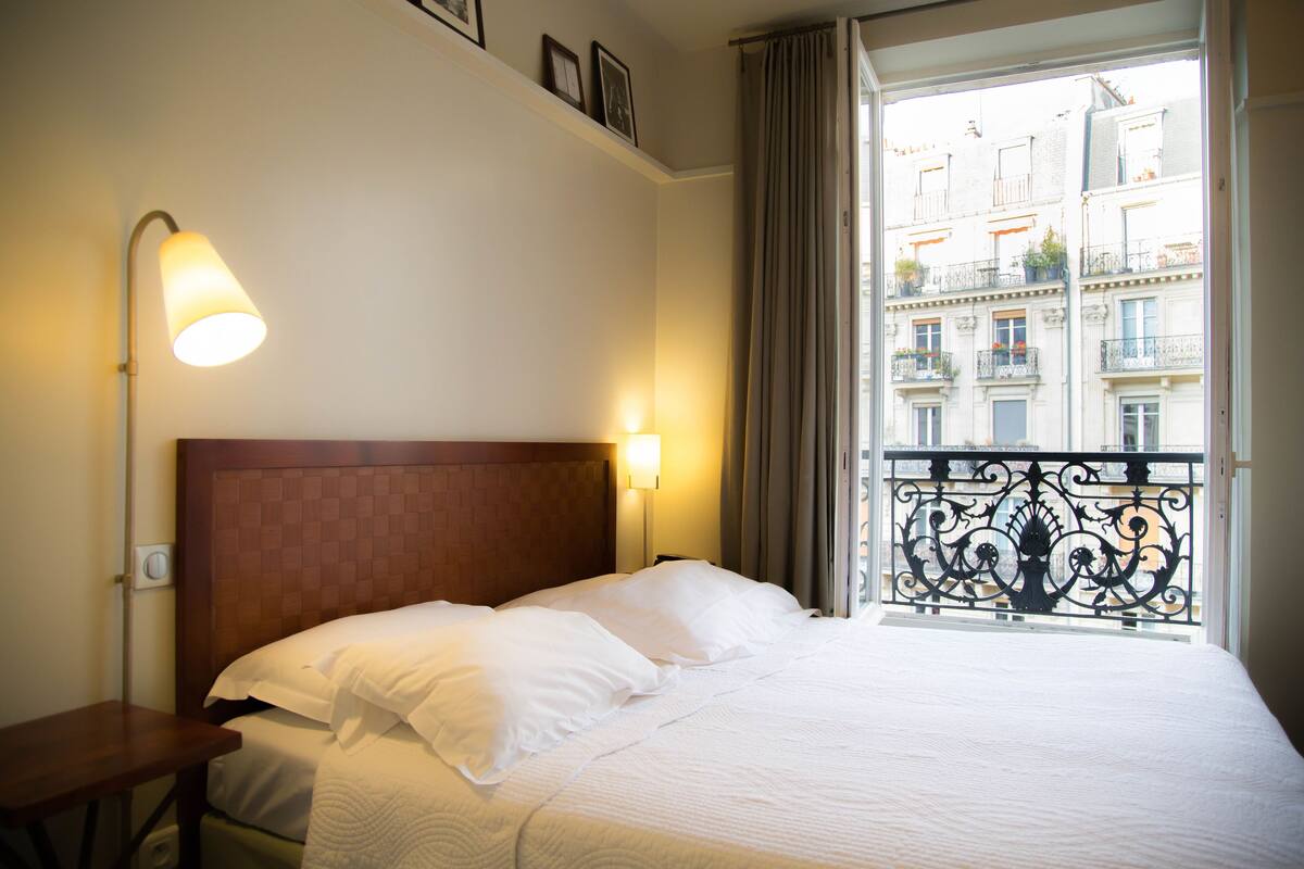 Quarto de hotel com cama, duas luminárias de cabeceira, janela com vista para edifícios e cortina.