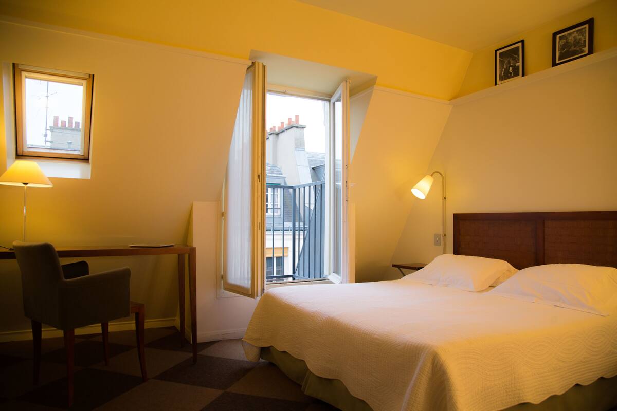 Quarto de hotel com cama, escrivaninha, cadeira, janela e porta para uma varanda.