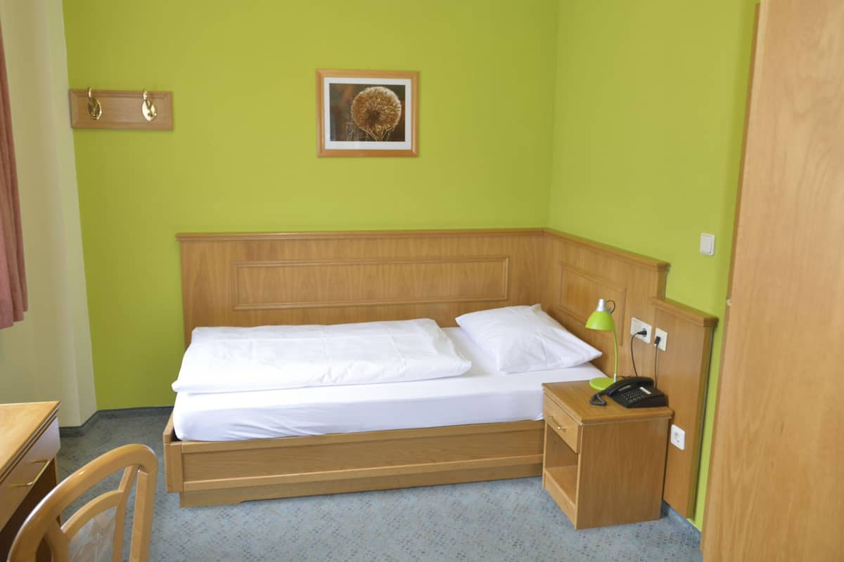 Uma cama de solteiro com cabeceira de madeira, uma mesinha de cabeceira com abajur e um criado-mudo de madeira.