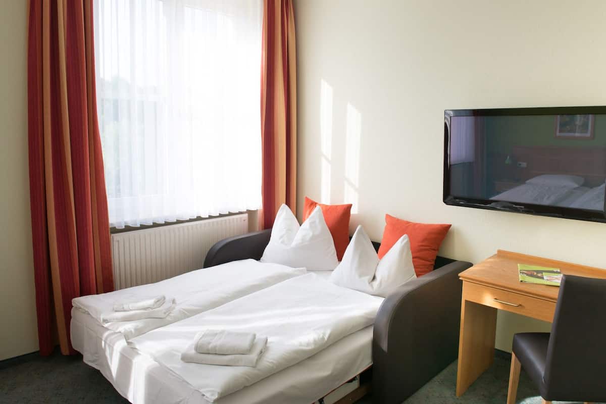 Quarto de hotel com cama, duas almofadas, escrivaninha, cadeira, televisão e janela com cortinas.