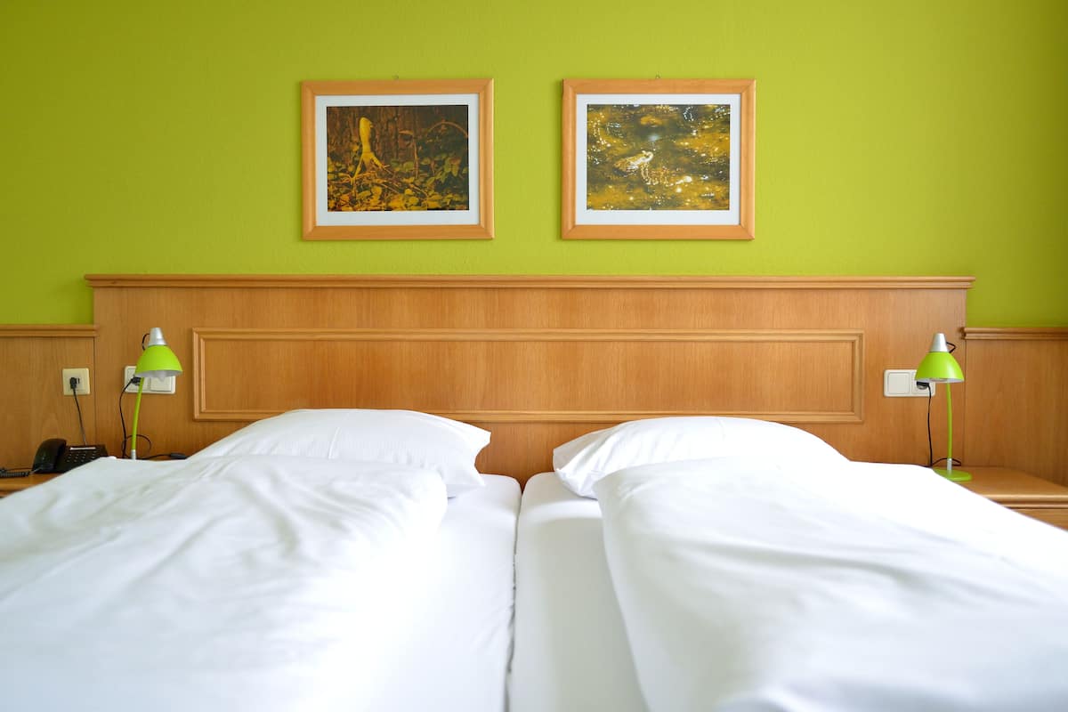 Quarto de hotel com duas camas, paredes verdes, cabeceira de madeira, quadros emoldurados e abajures de cabeceira.