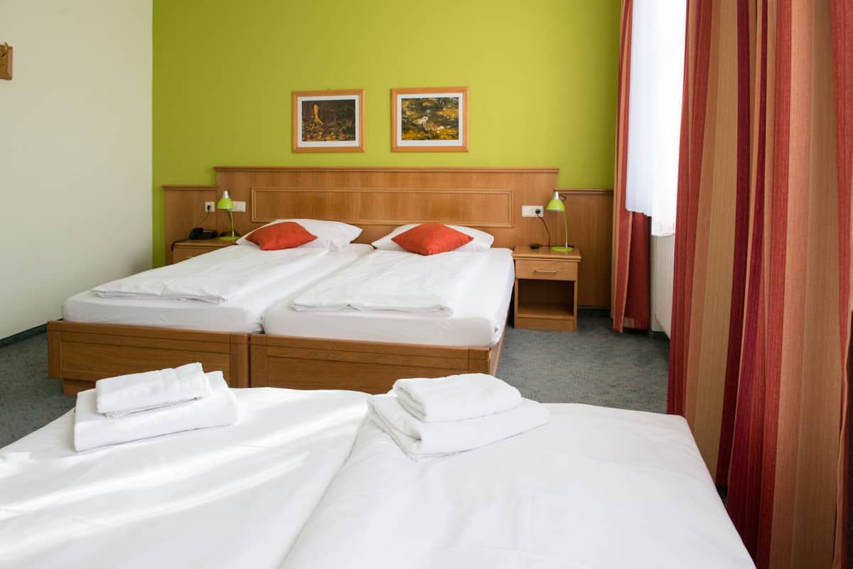 Quarto de hotel com duas camas, uma cabeceira de madeira, duas molduras na parede e uma mesa de cabeceira com abajur.
