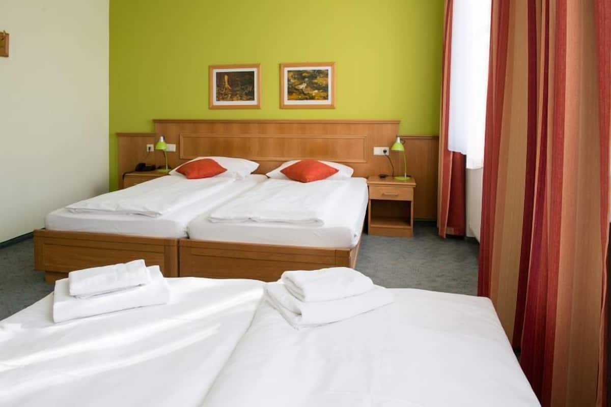 Uma cama de casal com lençóis brancos e almofadas vermelhas, uma cabeceira de madeira, duas mesas de cabeceira com abajures e duas fotos emolduradas na parede.