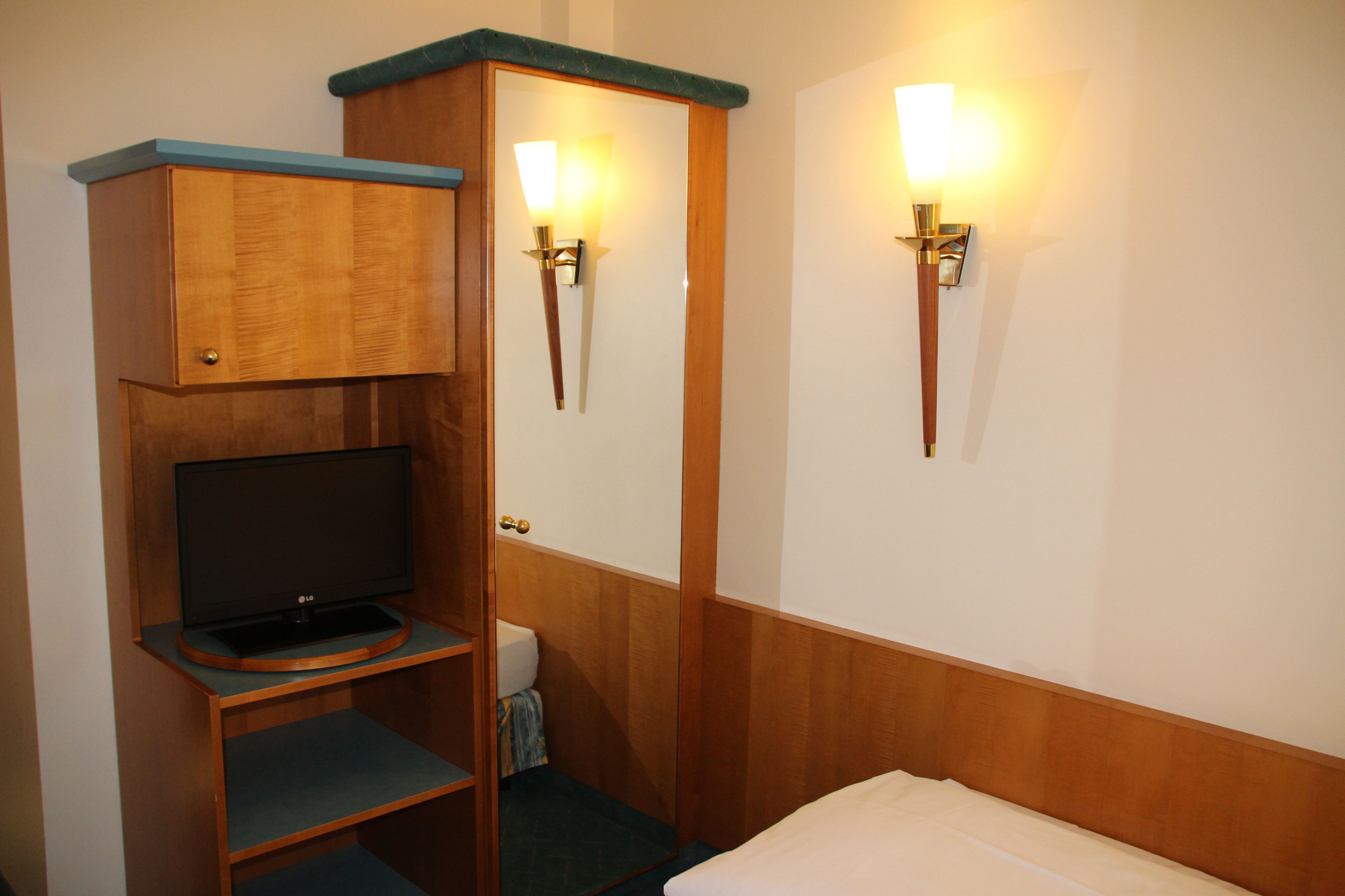 Quarto de hotel pequeno com um armário de madeira, uma televisão sobre um suporte, uma cama e duas luzes fixadas na parede.