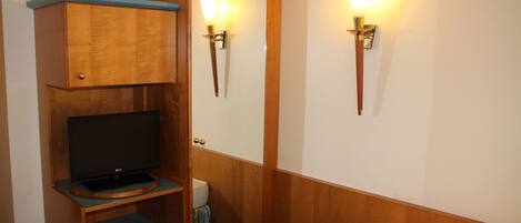 Quarto de hotel pequeno com um armário de madeira, uma televisão sobre um suporte, uma cama e duas luzes fixadas na parede.