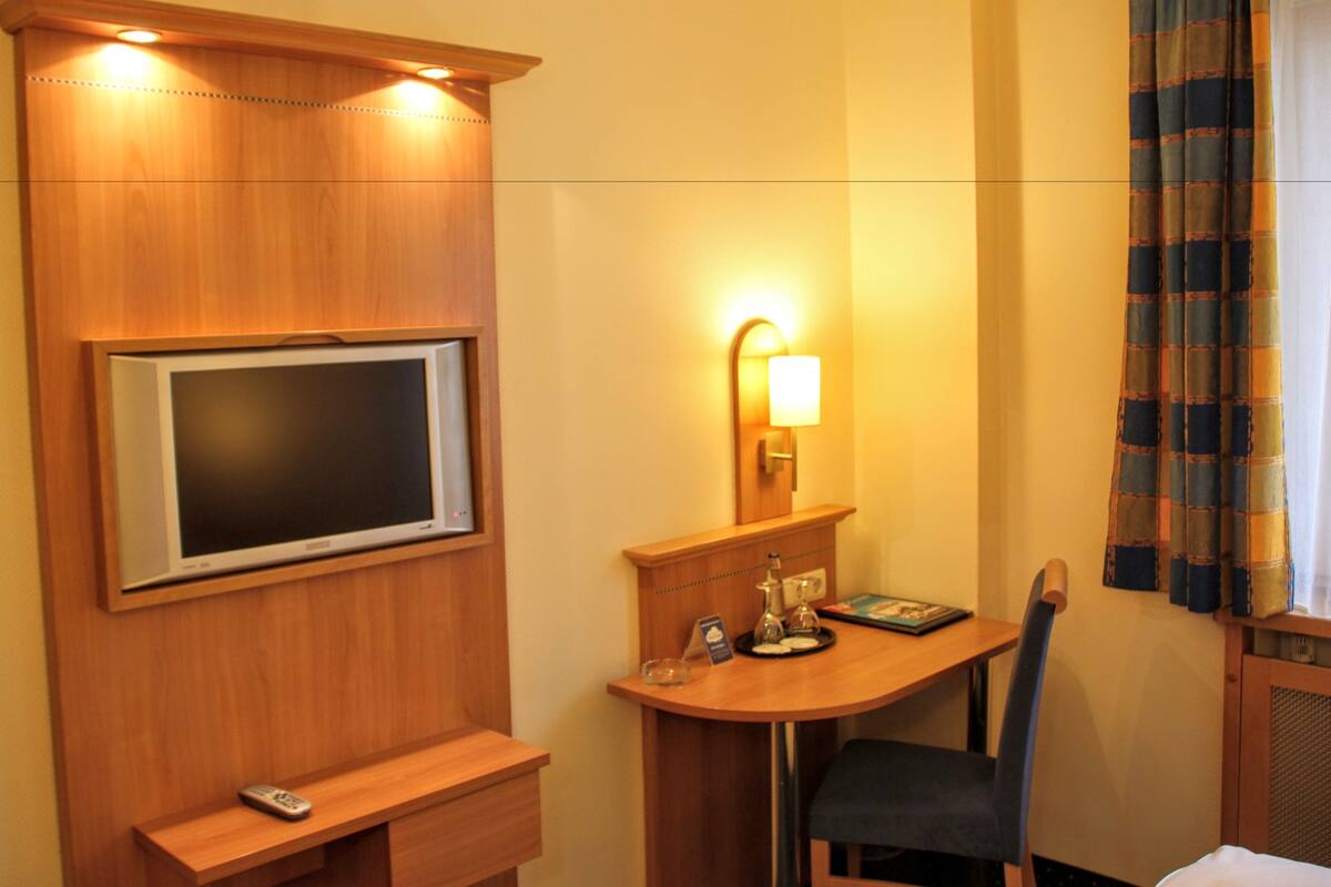 Quarto de hotel com um suporte de televisão de madeira, uma escrivaninha com cadeira, mesa de cabeceira e uma luminária.