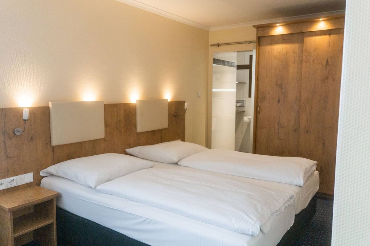 Quarto de hotel com cama de casal, cabeceira de madeira, mesas de cabeceira e guarda-roupa de madeira.