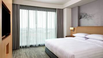 Quarto de hotel com uma cama grande, televisão de tela plana, luminária fixada na parede e janela com cortinas translúcidas.