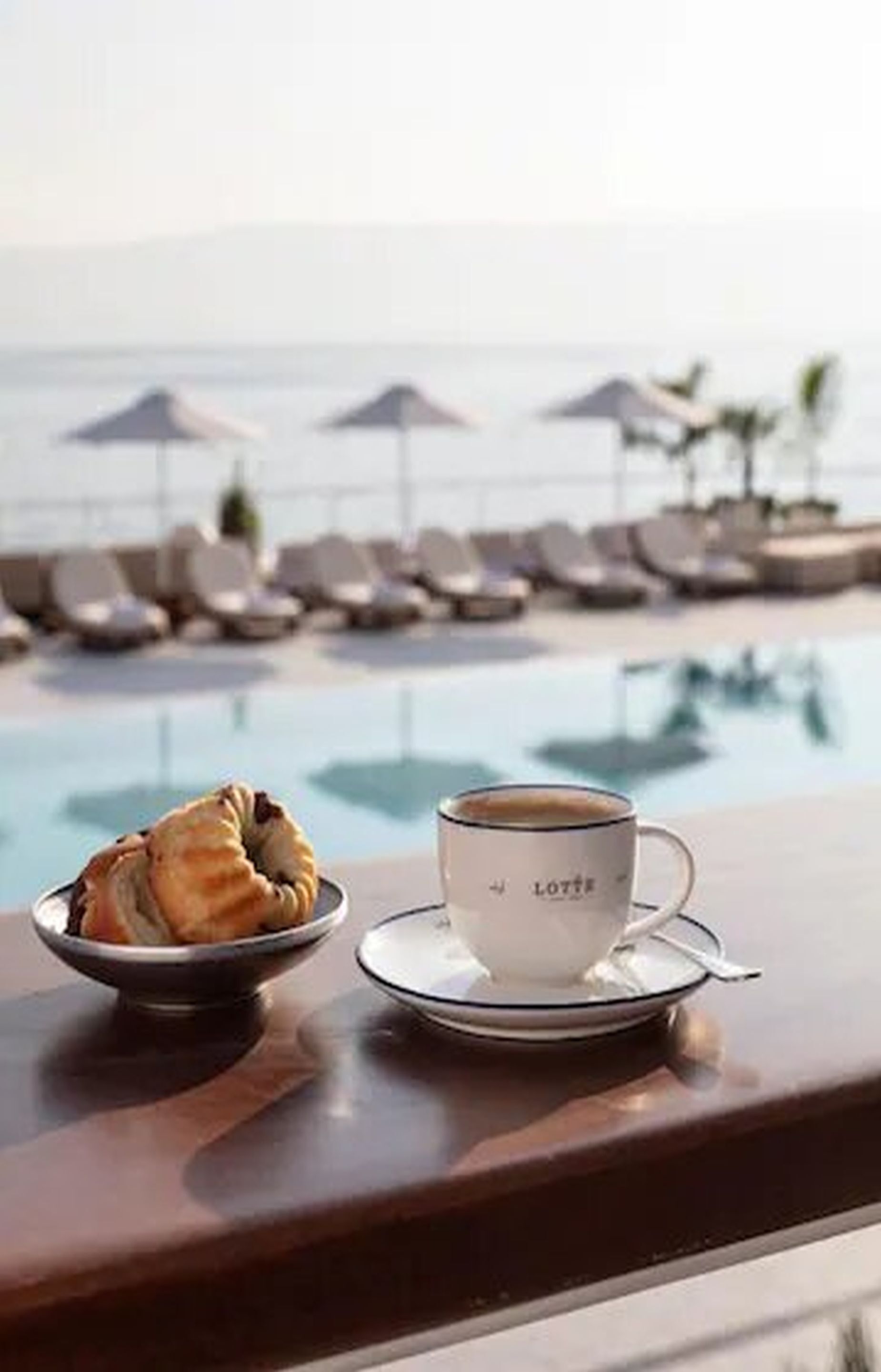 Mesa à beira da piscina com um croissant e uma chávena de café, com vista para a piscina e espreguiçadeiras com guarda-sóis.