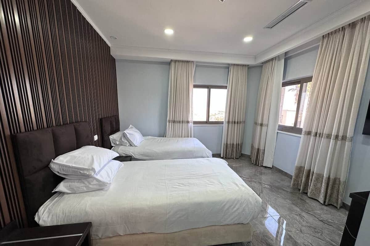 Quarto de hotel com duas camas, cabeceira de madeira, mesa de cabeceira e uma janela grande com cortinas.