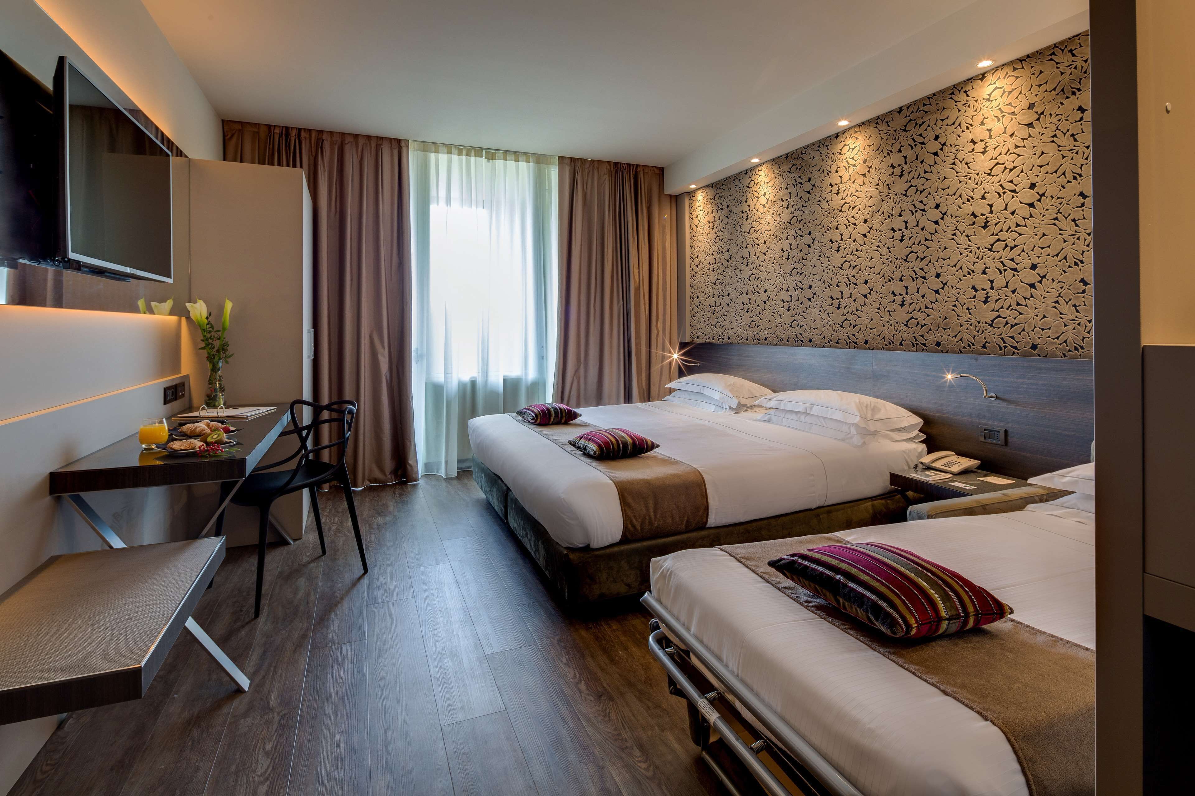 Quarto de hotel com duas camas, uma secretária com cadeira, televisão e uma janela grande com cortinas.