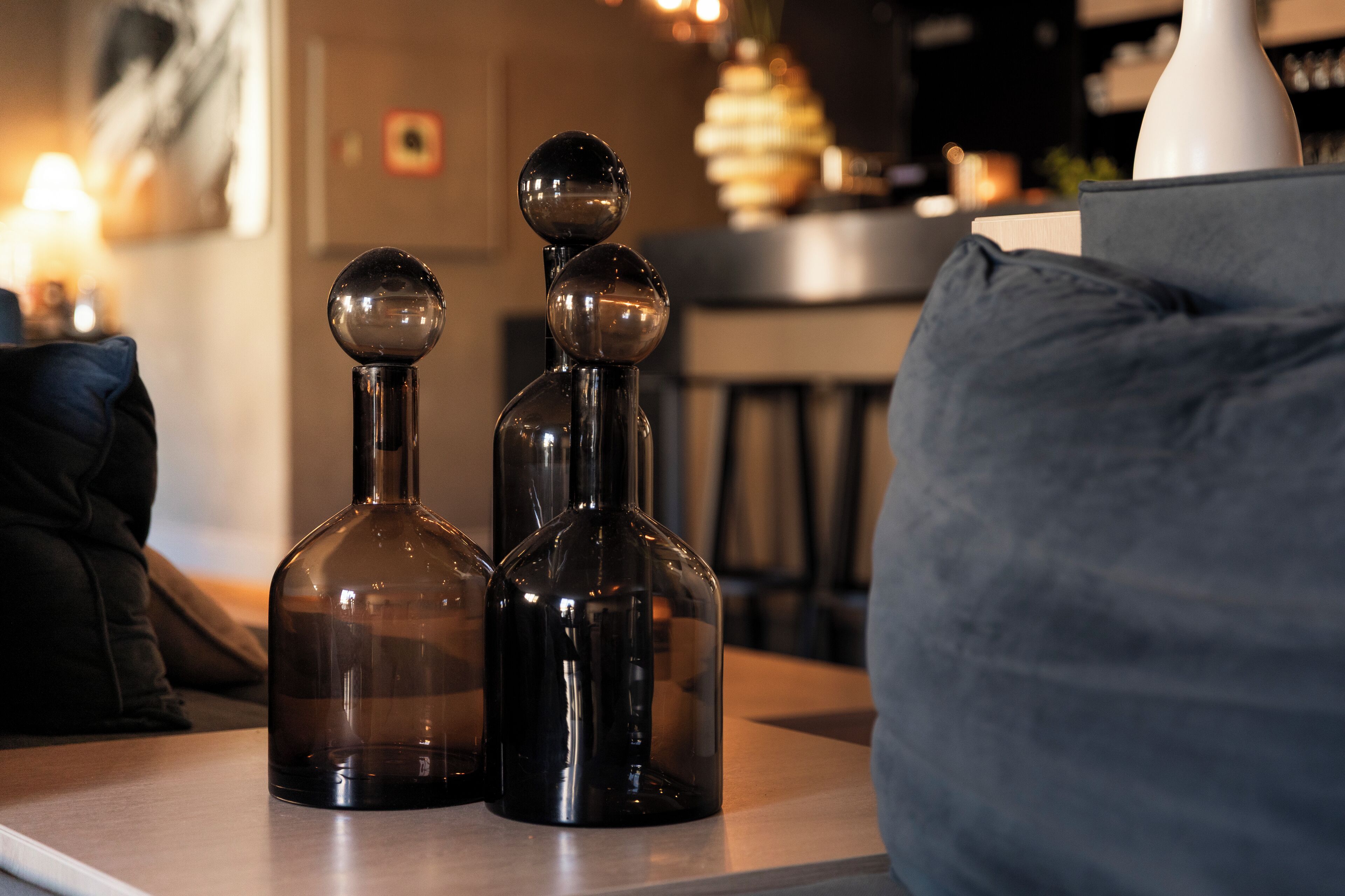 Três garrafas de vidro escuro sobre uma mesa em uma sala de estar moderna, com um sofá e uma luminária ao fundo.