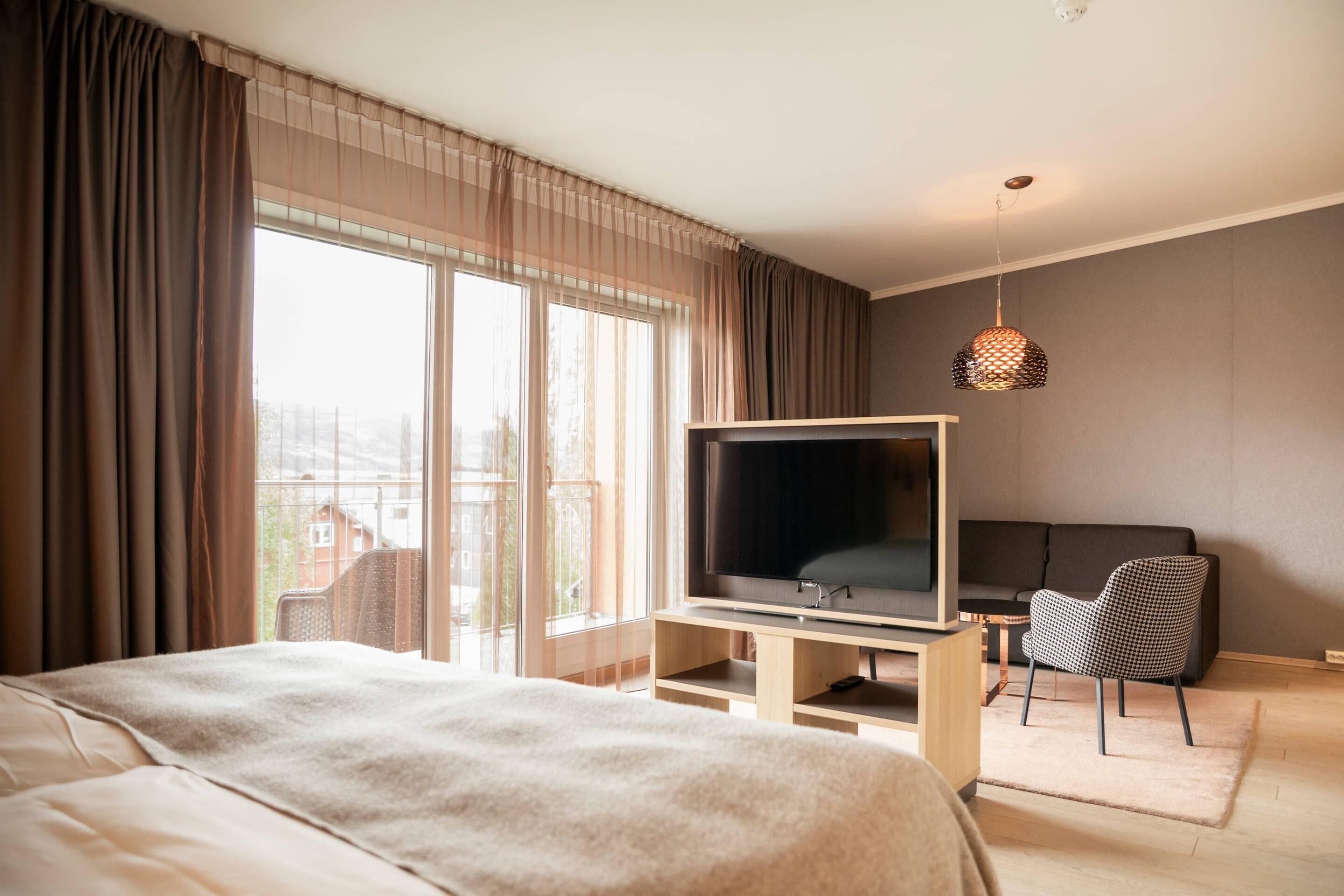 Quarto de hotel com uma cama grande, uma televisão sobre suporte de madeira, um sofá e uma varanda com cortinas.