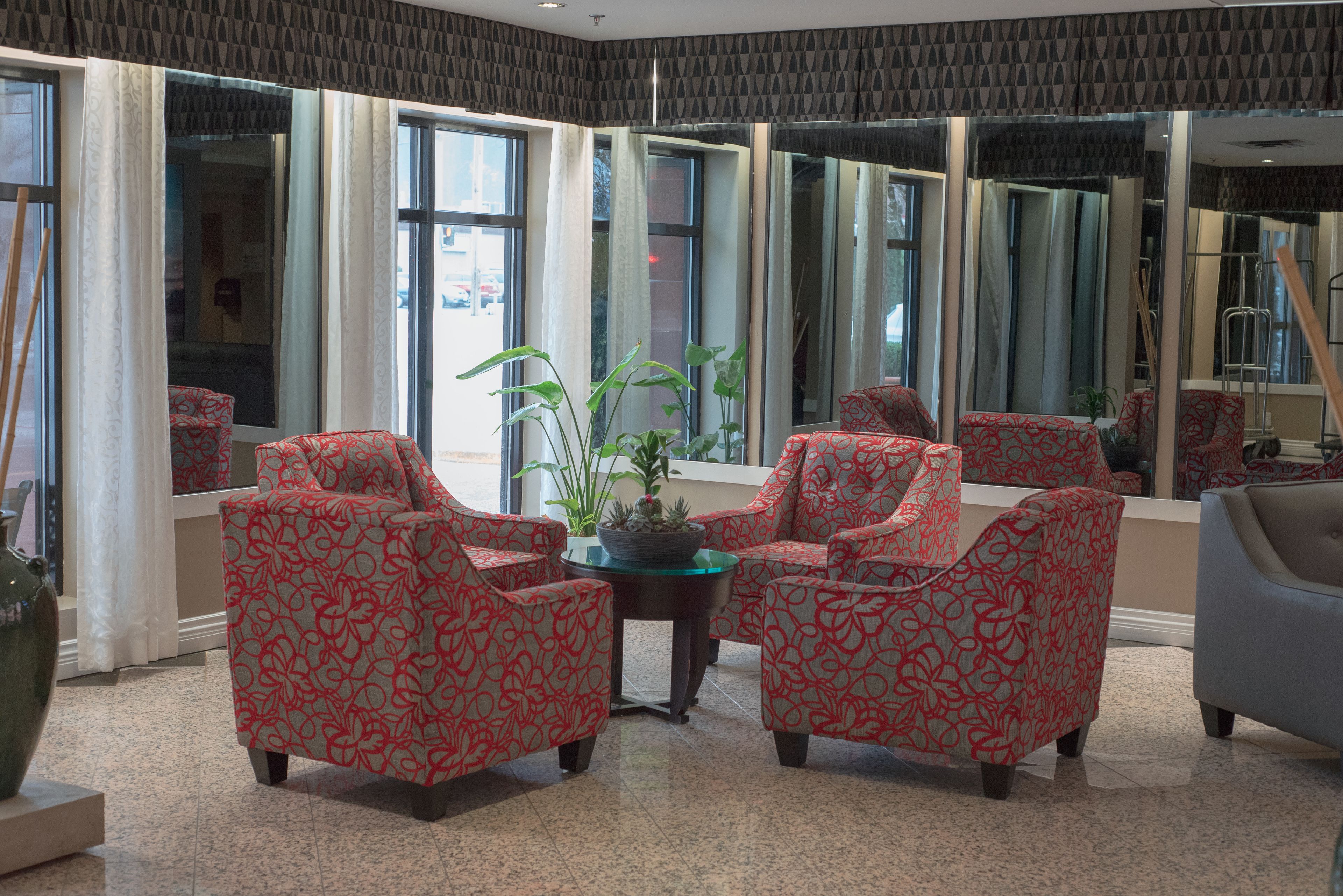Um hall com cadeiras estampadas em vermelho, uma mesa de vidro com uma planta e janelas amplas com cortinas.
