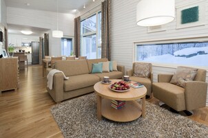 Uma sala de estar moderna com sofá, poltronas, uma mesa de centro com uma cesta de frutas e vista para uma paisagem nevada através de grandes janelas.