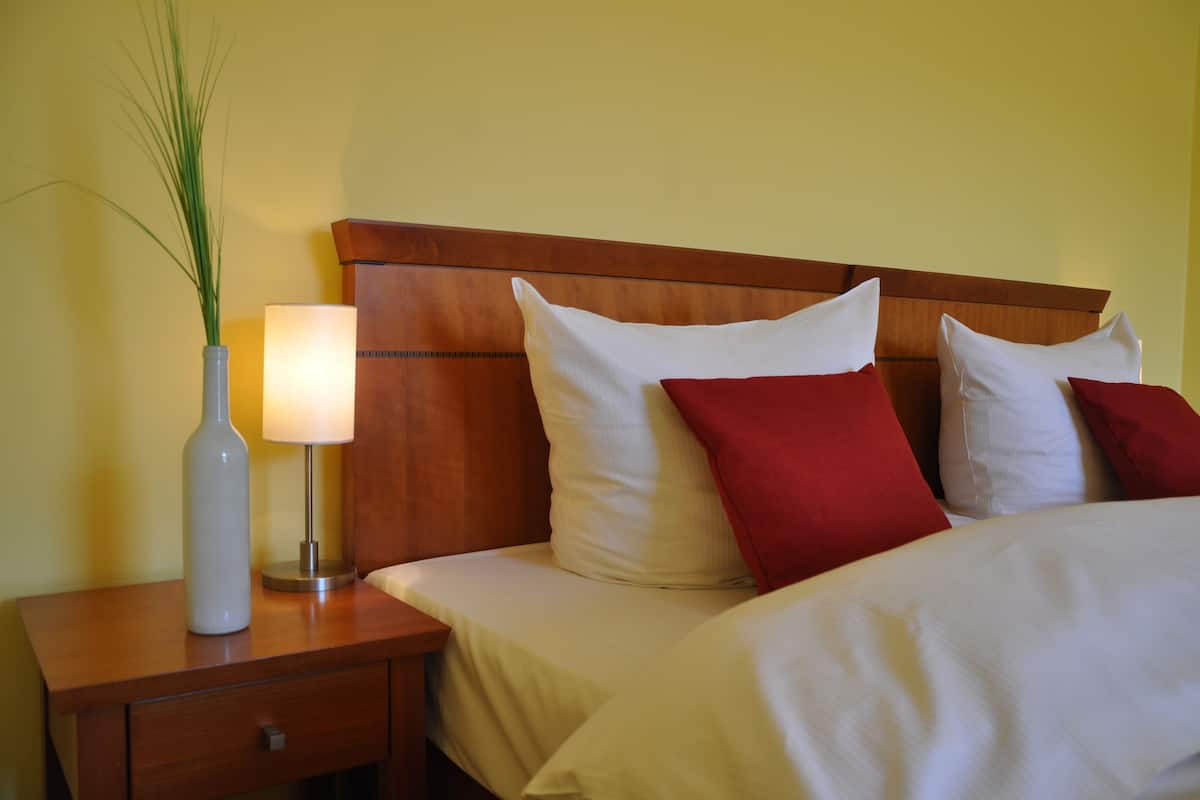 Cama bem arrumada com almofadas brancas e vermelhas, cabeceira de madeira e mesa de cabeceira com abajur e um vaso com folhagem.