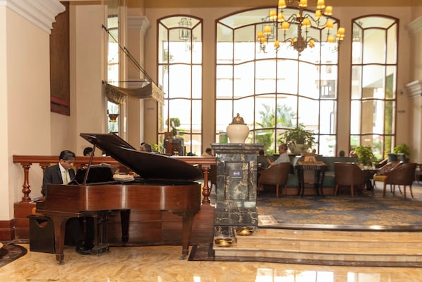 Um amplo hall com um piano de cauda, piso de mármore e grandes janelas em arco com um lustre.