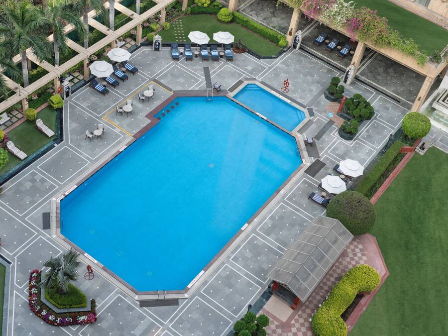 Uma grande piscina retangular com espreguiçadeiras e guarda-sóis, cercada por um espaço pavimentado com árvores e um pequeno jardim.