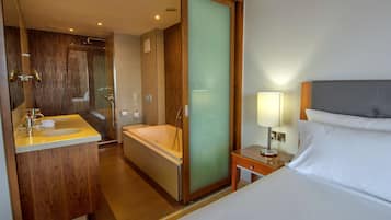Quarto de hotel com penteadeira de madeira, banheira, cama com roupa branca e mesa de cabeceira com abajur.