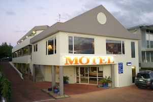 Um motel de dois andares com um letreiro grande 'MOTEL', uma escadaria que dá acesso ao andar superior e um estacionamento com carros.