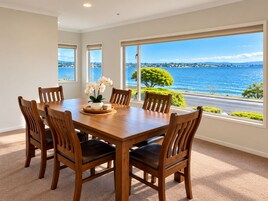 Uma sala de jantar com mesa e cadeiras de madeira, um vaso de flores e janelas amplas com vista para a água e vegetação.