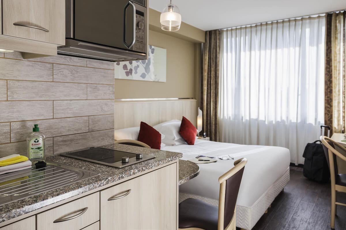 Quarto de hotel com cozinha compacta, cama com almofadas vermelhas, escrivaninha e vista para o exterior.