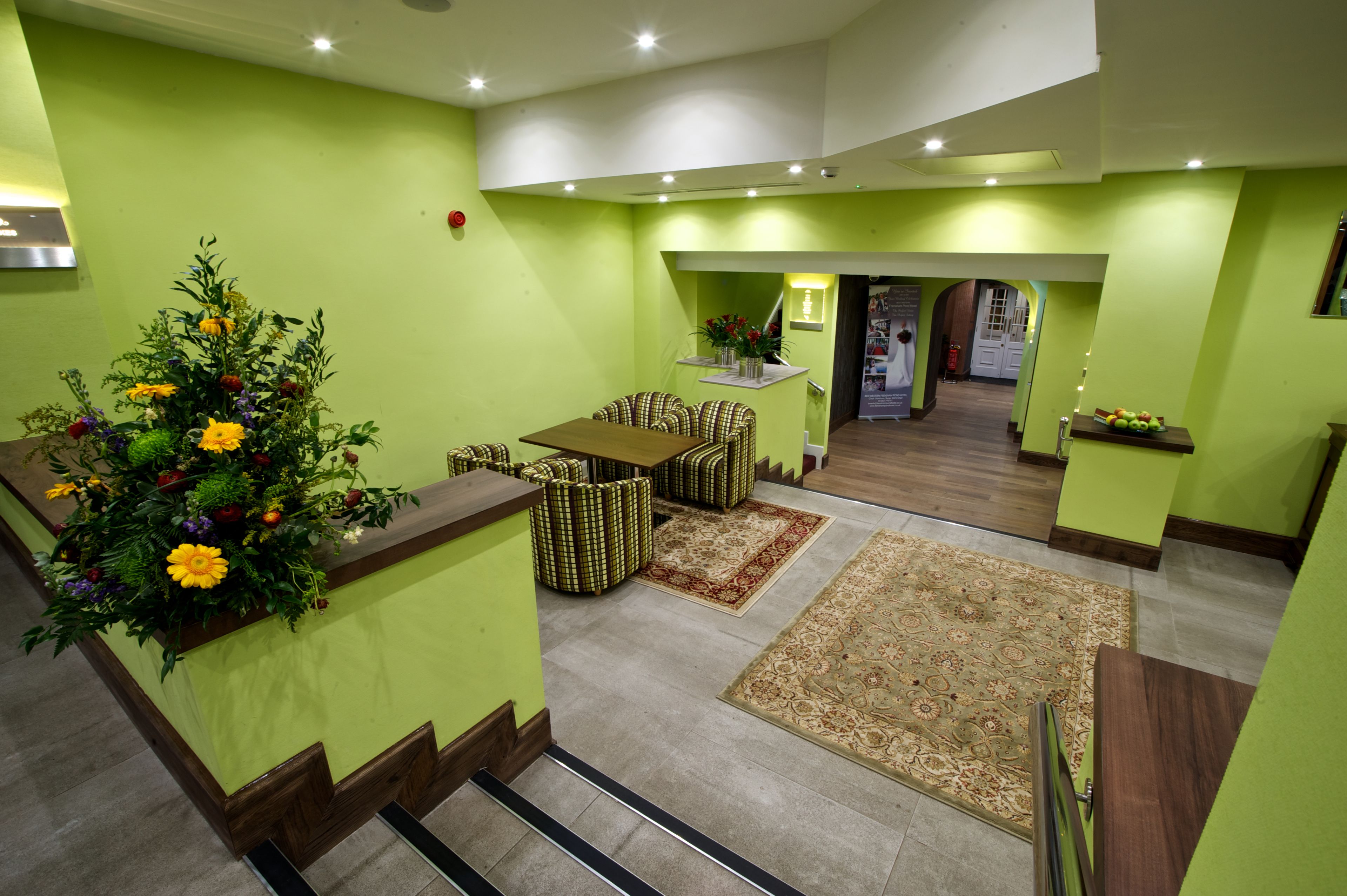 Um hall de hotel moderno com esquema de cores verde, balcão de recepção, área de estar com mesa e cadeiras, e um arranjo floral decorativo.