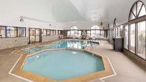 Uma piscina interior ampla com paredes de vidro transparente, borda em azulejo e área de estar com cadeiras e mesas.