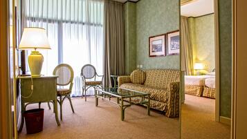 Quarto de hotel com parede verde, sofá estampado, mesa de centro de vidro, cadeira de vime, pequena mesa com abajur e cama com colcha listrada.