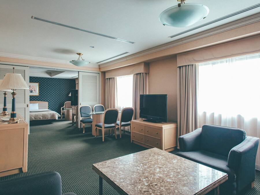 Quarto de hotel com cama, escrivaninha, cadeiras, televisão e uma grande janela com cortinas.