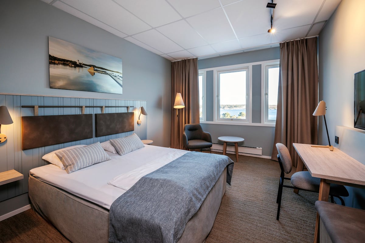 Quarto de hotel com uma cama grande, uma escrivaninha, uma cadeira, uma mesa pequena e um quadro na parede.