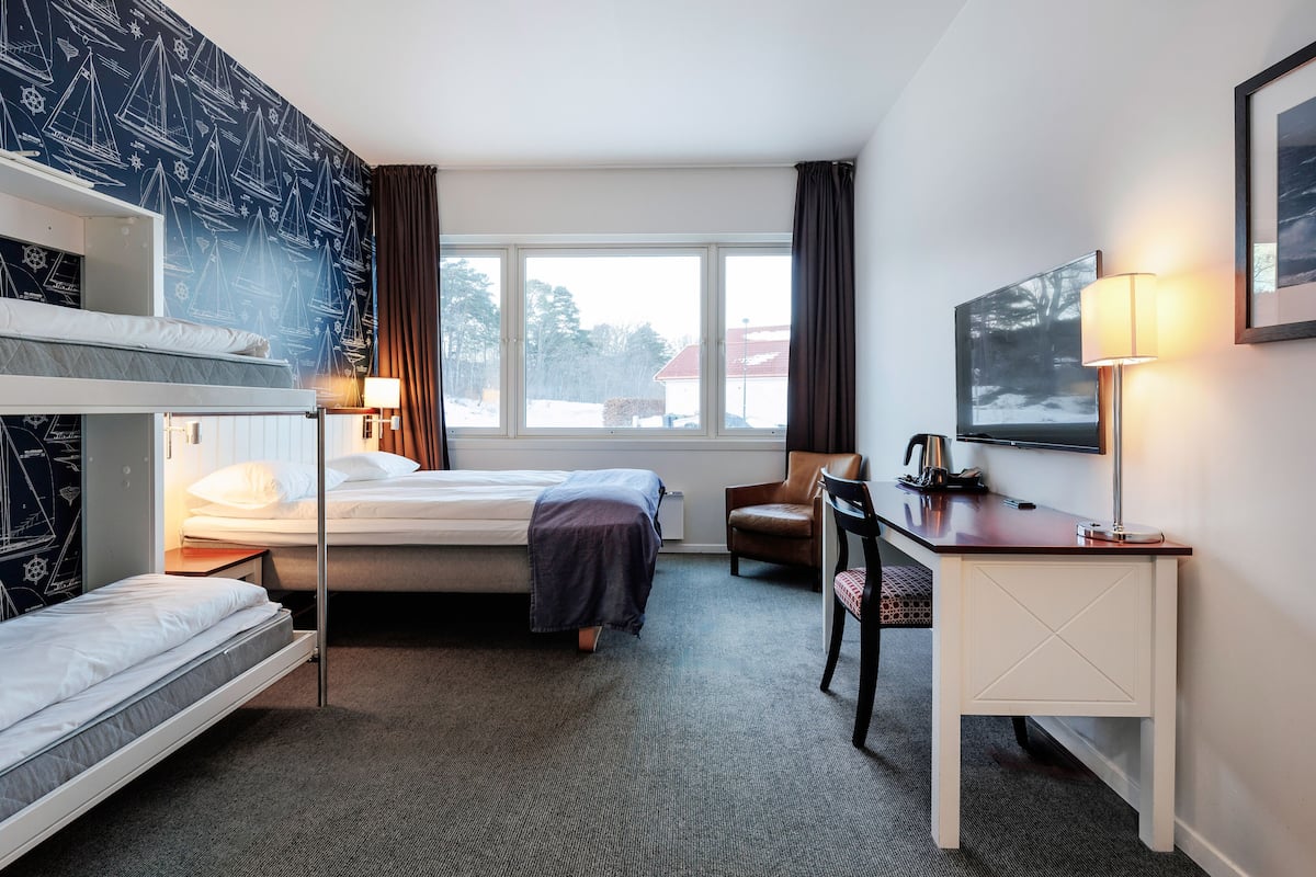 Quarto de hotel com cama de solteiro, uma cama individual, uma escrivaninha com cadeira, televisão e janela com cortinas.