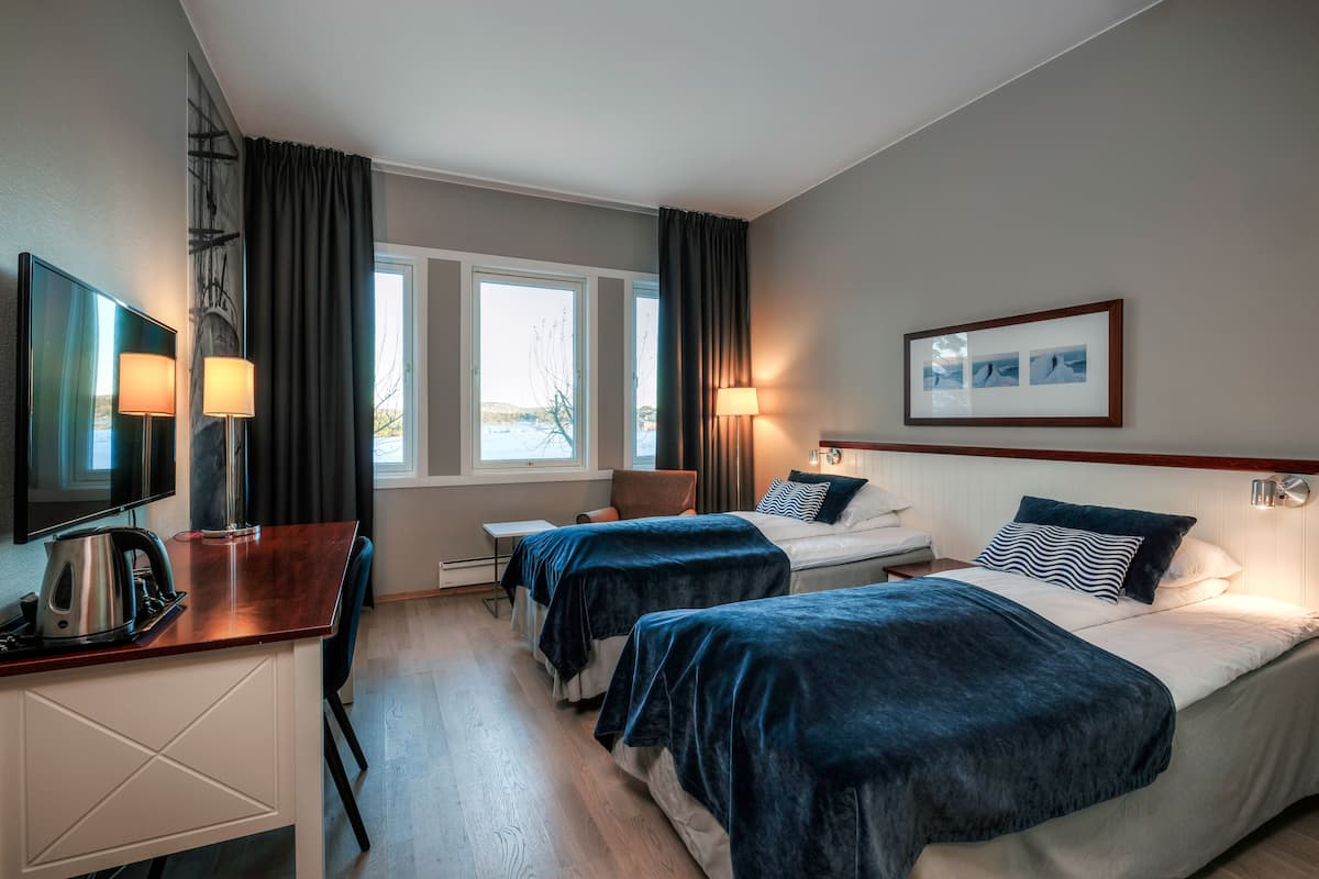 Quarto de hotel com duas camas, uma escrivaninha com chaleira, uma cadeira e uma televisão.