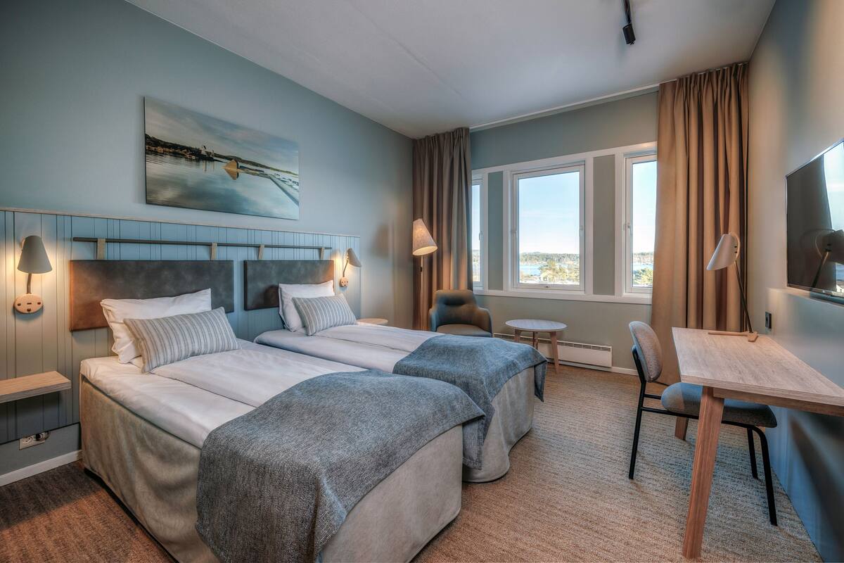 Quarto de hotel com duas camas, uma escrivaninha, uma cadeira e uma janela grande.