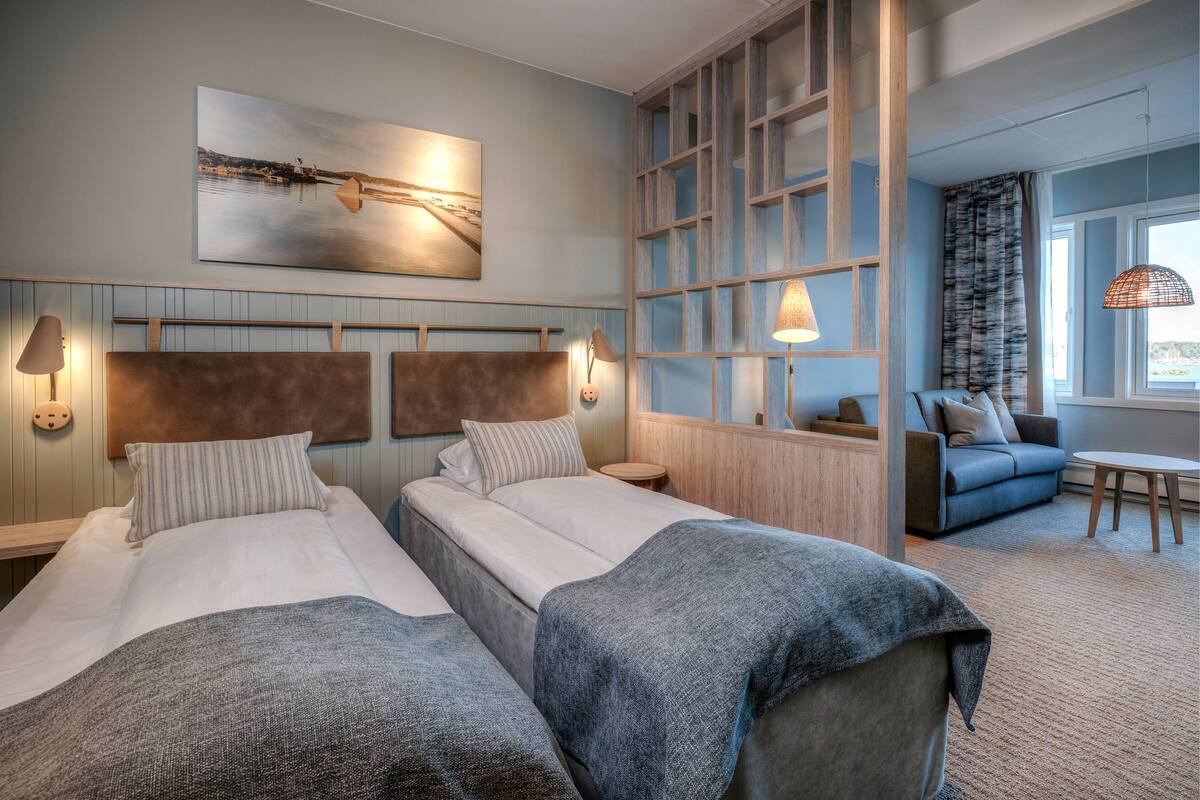 Quarto de hotel com duas camas, área de estar com sofá e poltronas, divisória de madeira e uma janela grande com vista.