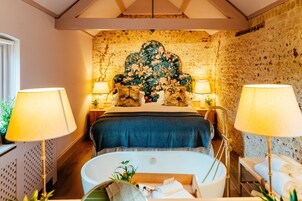 Um quarto com uma cama grande, duas luminárias de cabeceira, um painel de cabeceira de madeira, uma banheira e uma mesinha com uma planta.