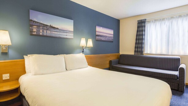Quarto de hotel com uma cama grande, um sofá, duas luminárias fixadas na parede e duas pinturas.