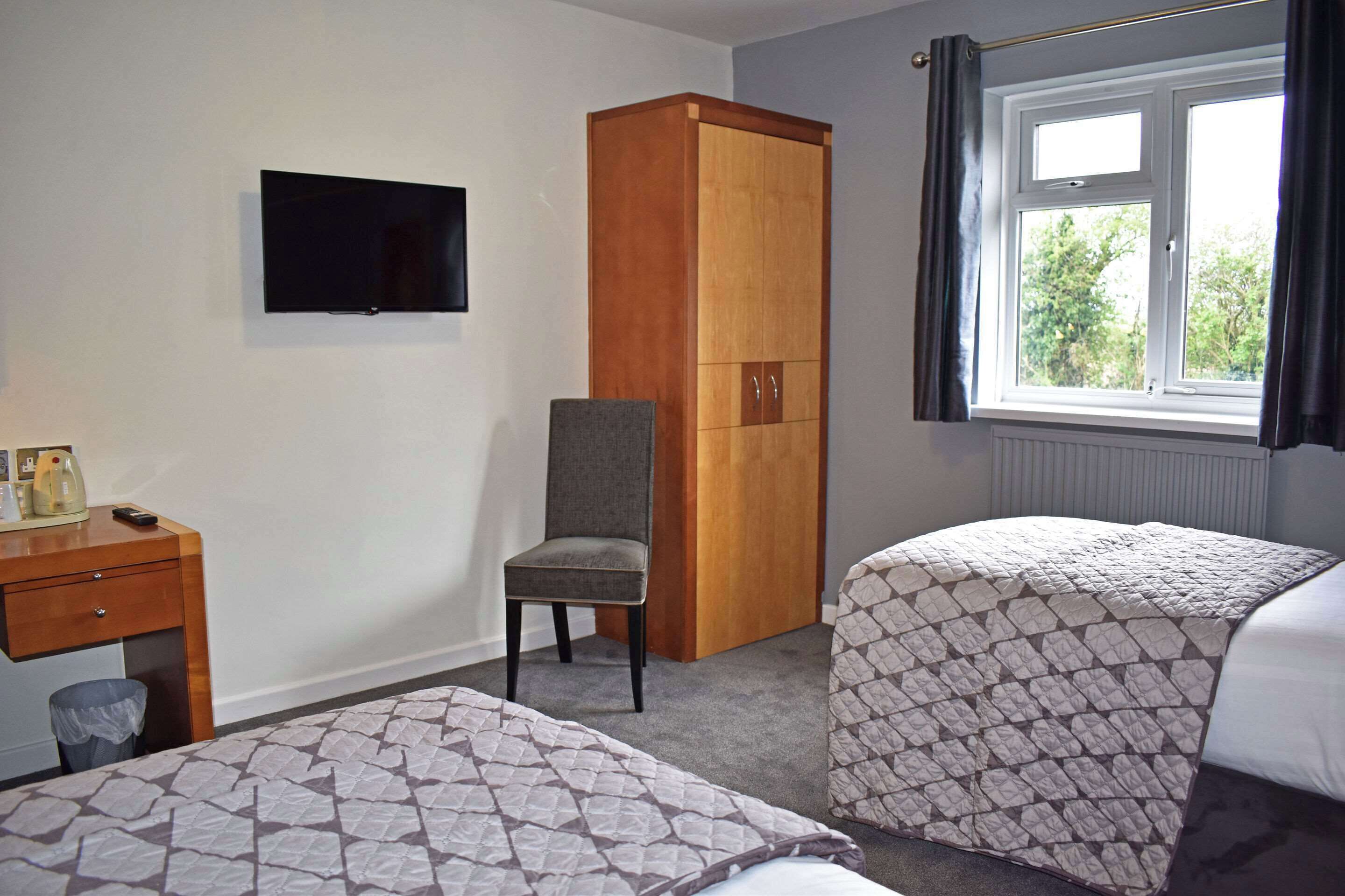Quarto de hotel com duas camas, televisão, guarda-roupa, cadeira e janela com cortinas.