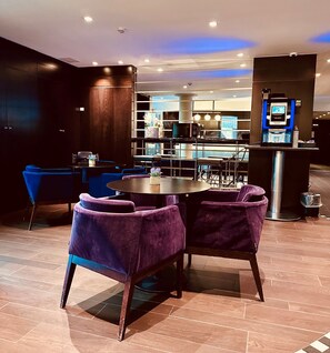 Um hall de hotel moderno com uma mesa redonda e cadeiras roxas, uma área de bar com uma máquina de café, e vista da cidade através de grandes janelas.