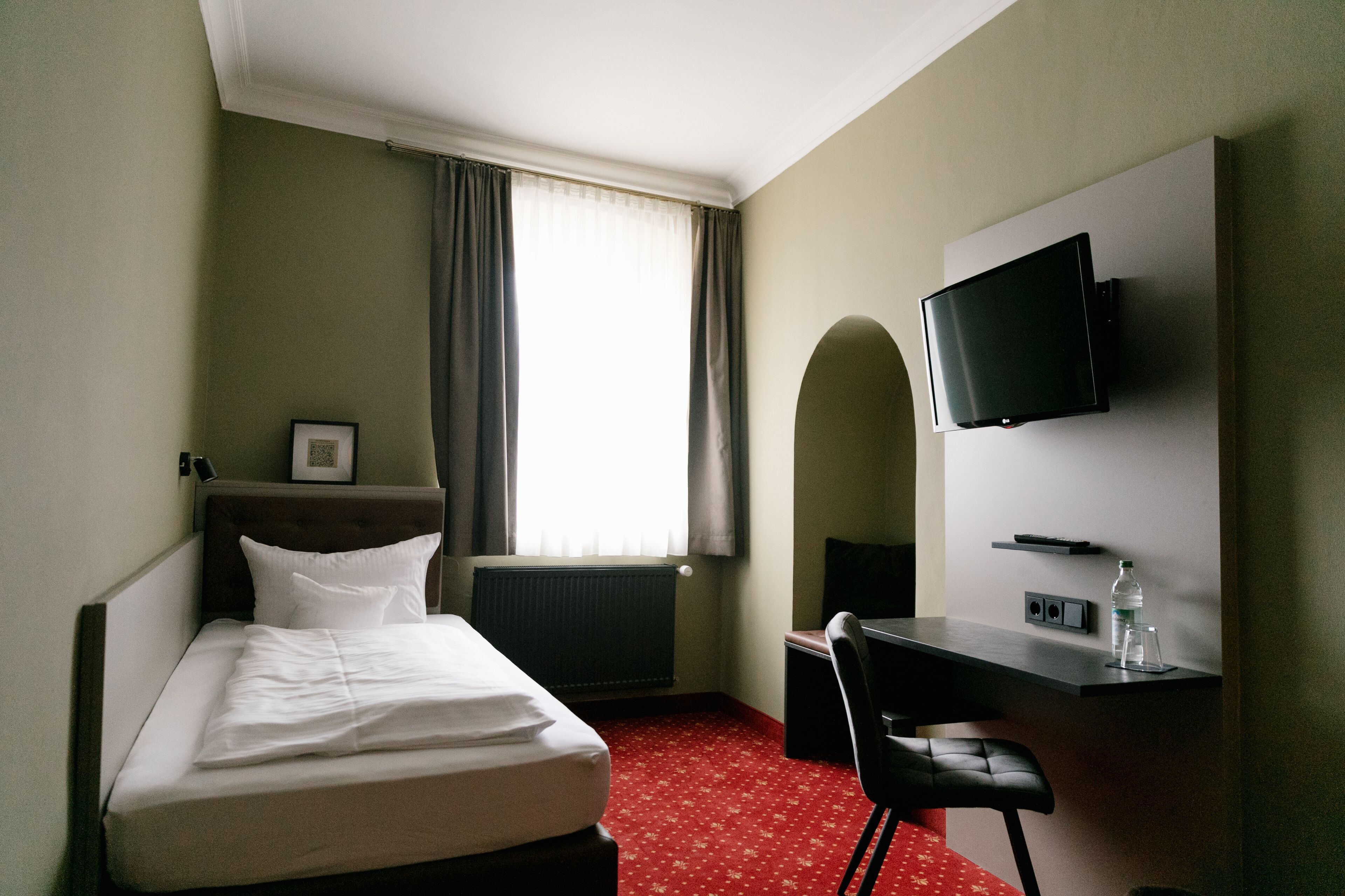 Quarto de hotel com cama, escrivaninha, cadeira, televisão e janela com cortinas.