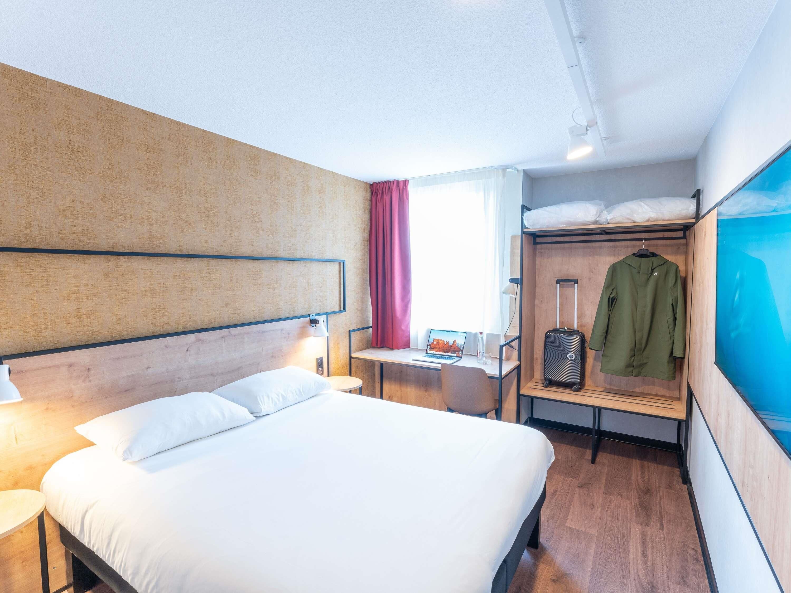 Quarto de hotel com uma cama, uma escrivaninha com um laptop, um cabideiro e uma janela com cortinas.