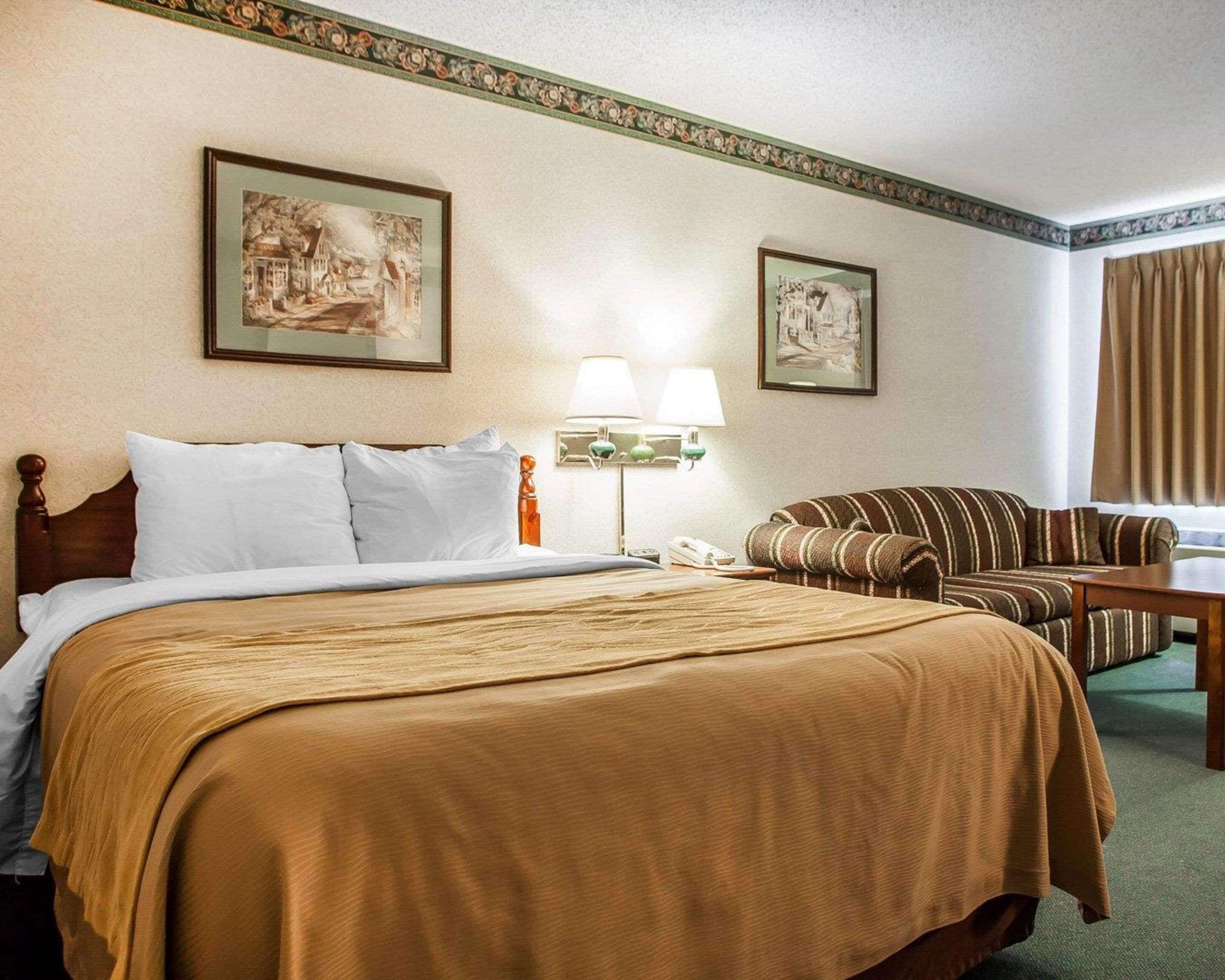 Quarto de hotel com uma cama grande, uma poltrona listrada, duas luminárias fixadas na parede e duas quadros emoldurados.