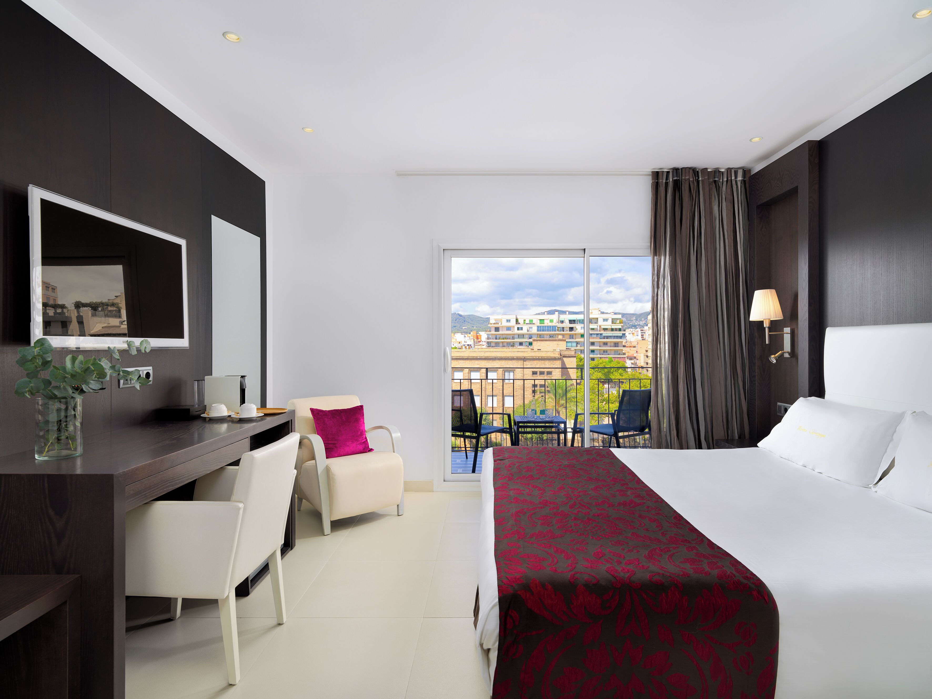 Quarto de hotel moderno com uma cama grande, uma escrivaninha com cadeira, uma mesa pequena e uma varanda com vista para a cidade.