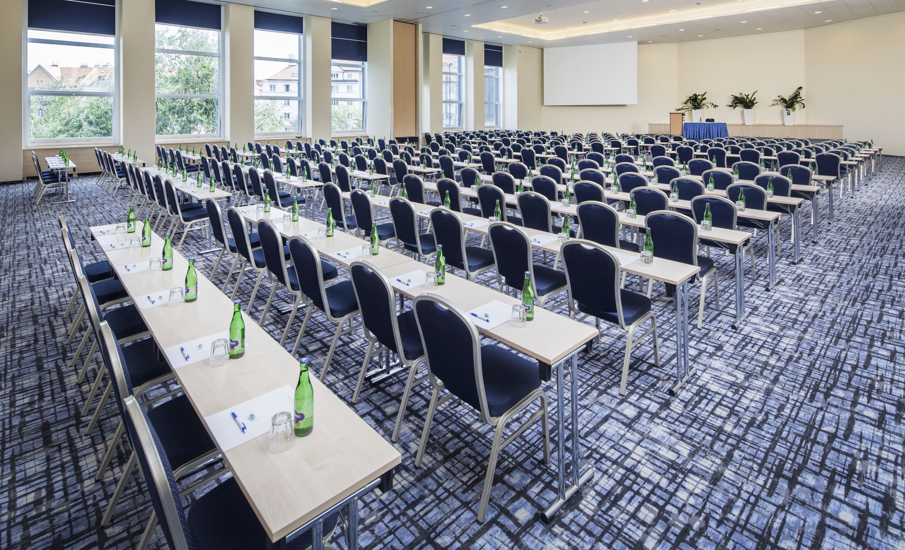 Uma sala de conferências com fileiras de mesas e cadeiras, janelas amplas e uma tela de projeção.