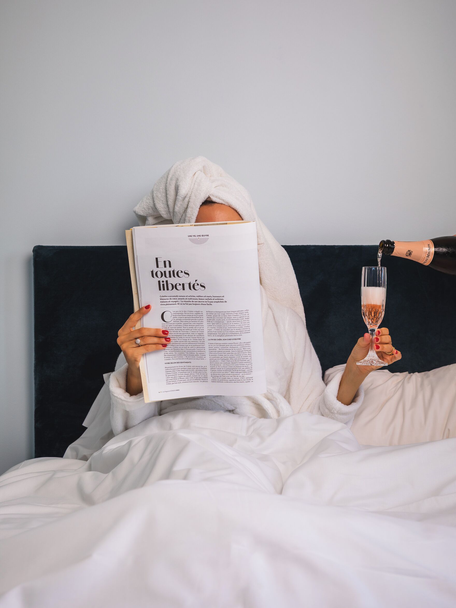 Uma pessoa lendo um livro na cama enquanto champanhe é servido em uma taça.