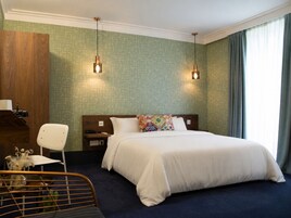 Quarto de hotel com uma cama grande, duas luminárias de cabeceira, uma escrivaninha de madeira, uma cadeira e uma janela com cortinas.