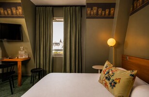 Quarto de hotel com cama, escrivaninha, cadeira, televisão e janela com cortinas.